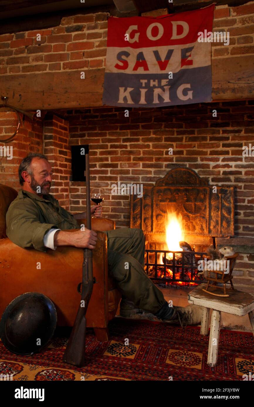 Man in green overalls seated by roaring fire with rifle and red wine ...