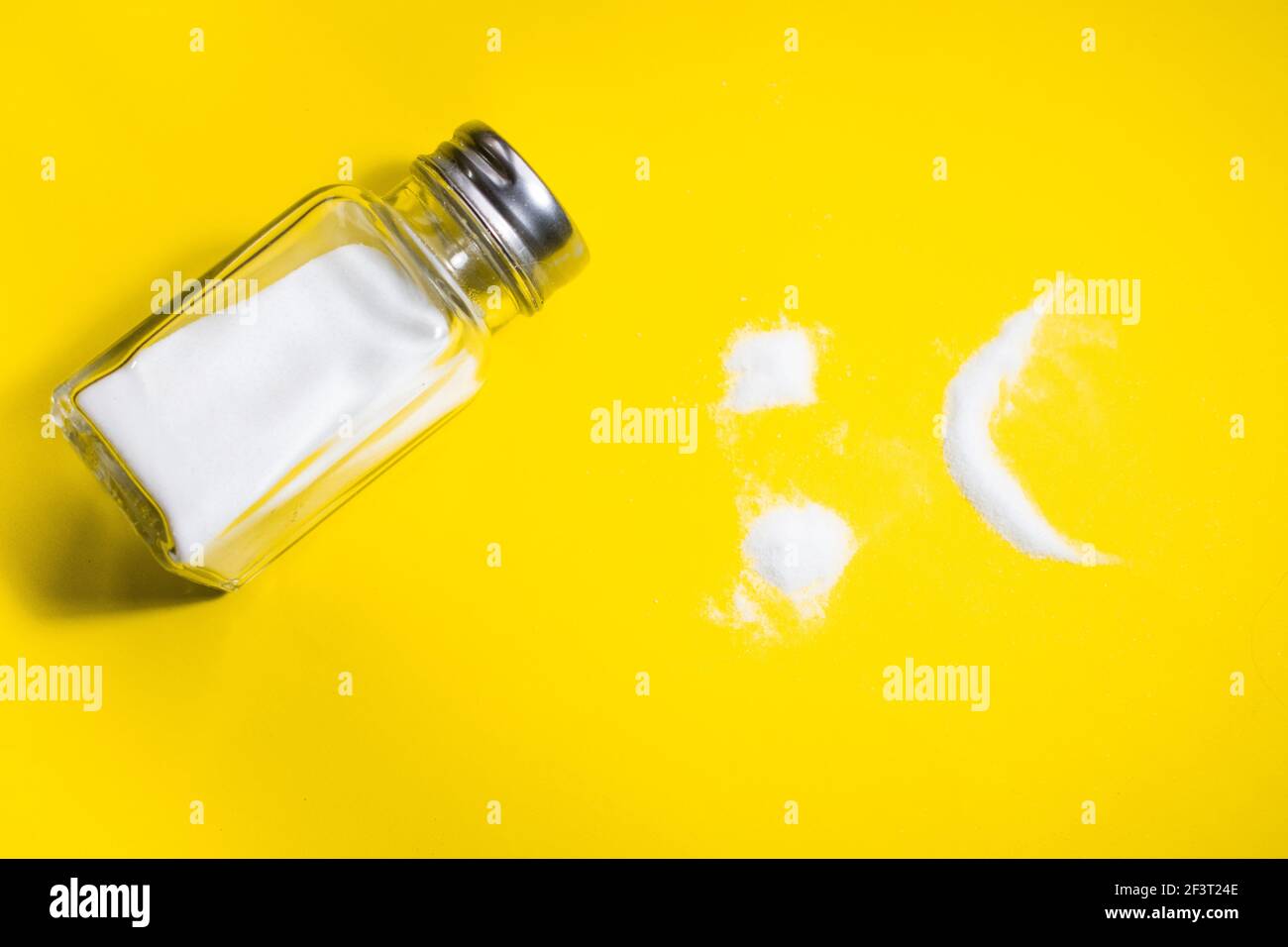A top view of a sad face made of salt and a glass shaker on a yellow ...