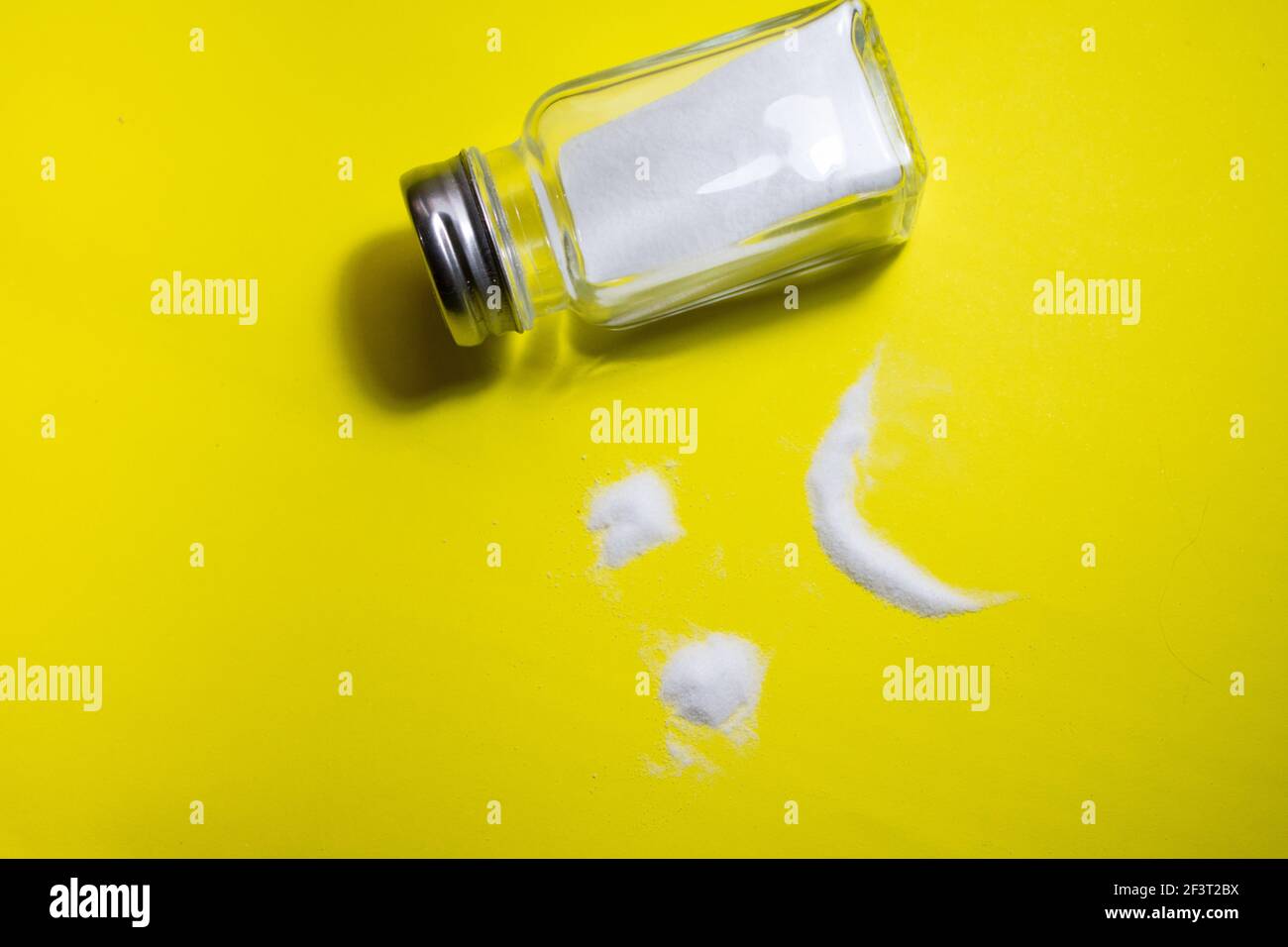 A top view of a sad face made of salt and a glass shaker on a yellow ...