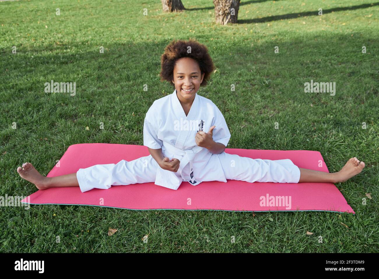 Cheerful little girl karateka sitting on yoga mat and giving thumbs up ...
