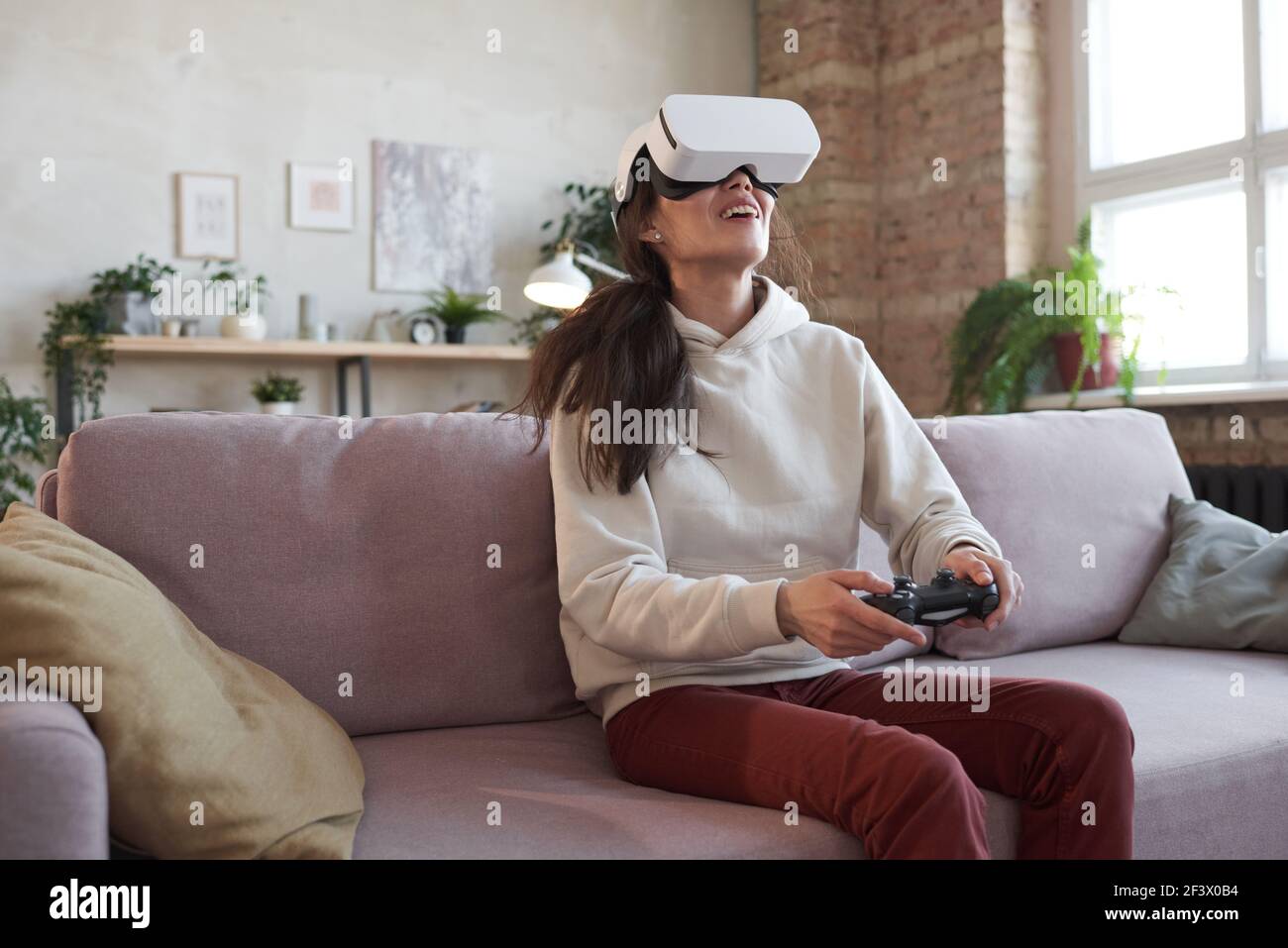 Young woman in vr glasses sitting on sofa and using joystick during her ...