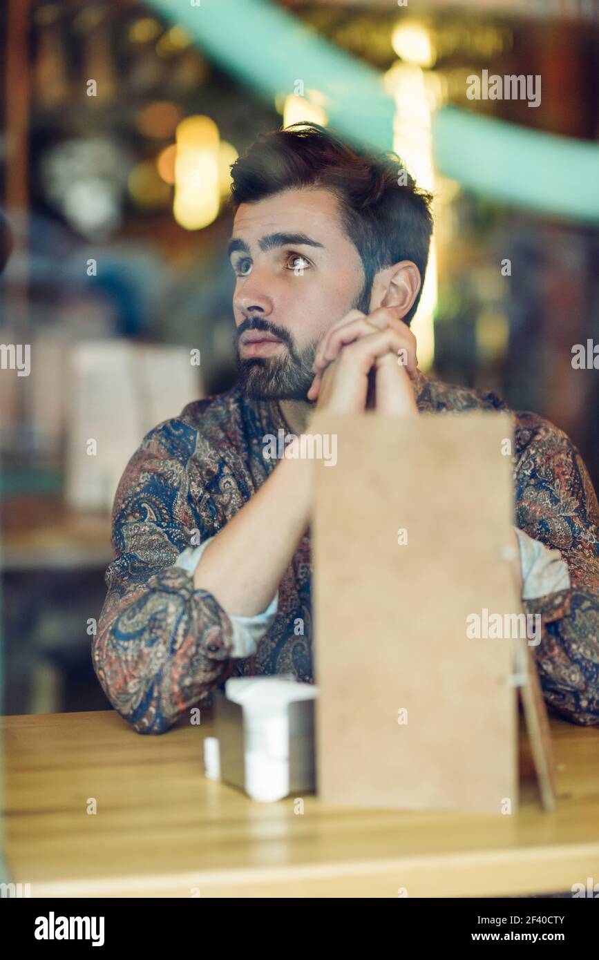 Thoughtful bearded man wearing casual clothes sitting in a modern pub ...