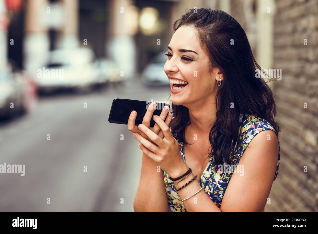 Smiling young woman recording voice note in her smart phone outdoors ...