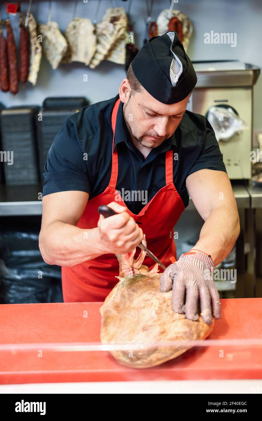 Male butcher boning a ham in a modern butcher shop with metal safety ...