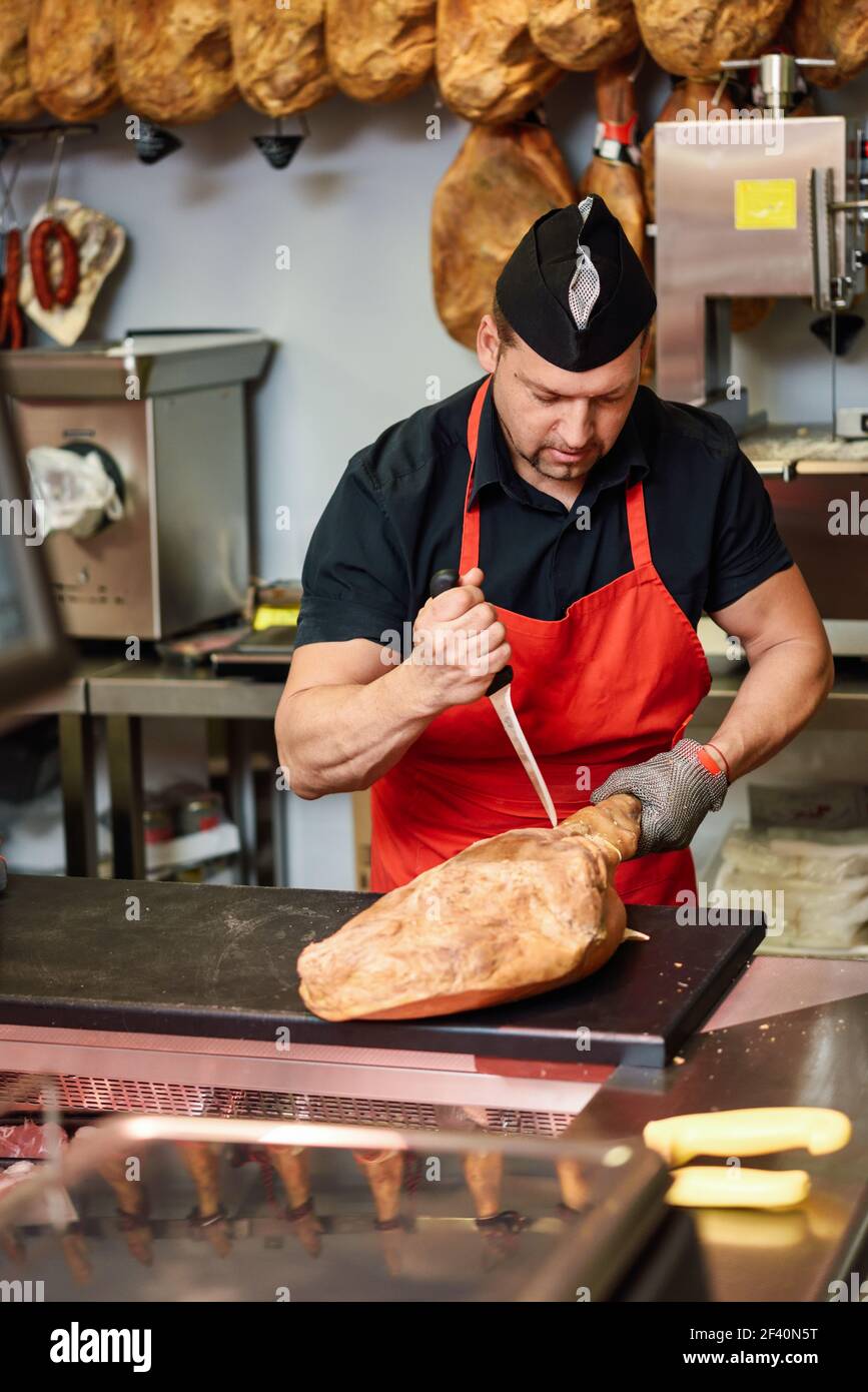 Male butcher boning a ham in a modern butcher shop with metal safety ...