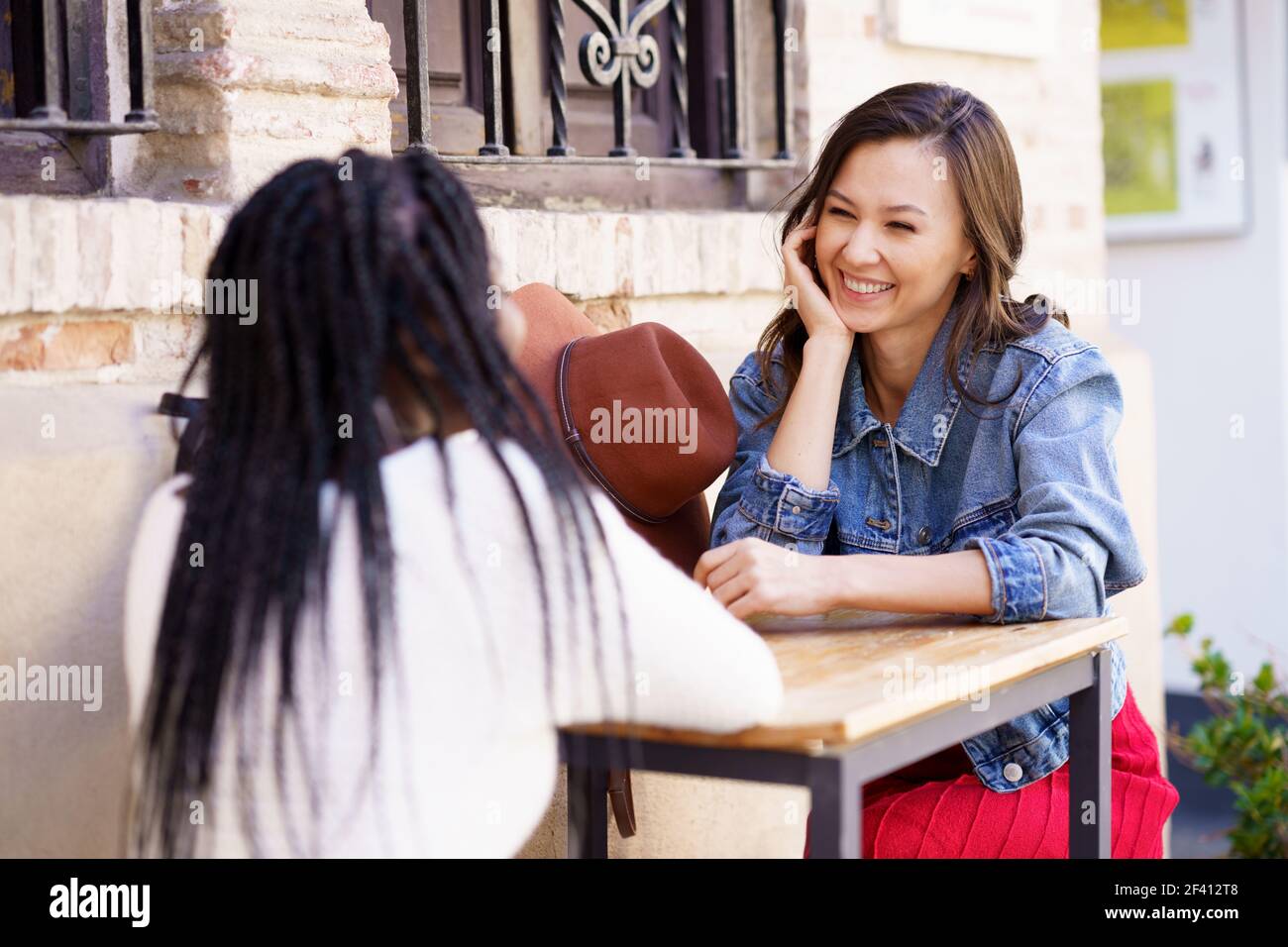 Two friends talking sitting at a table outside a bar. Multiethnic women ...