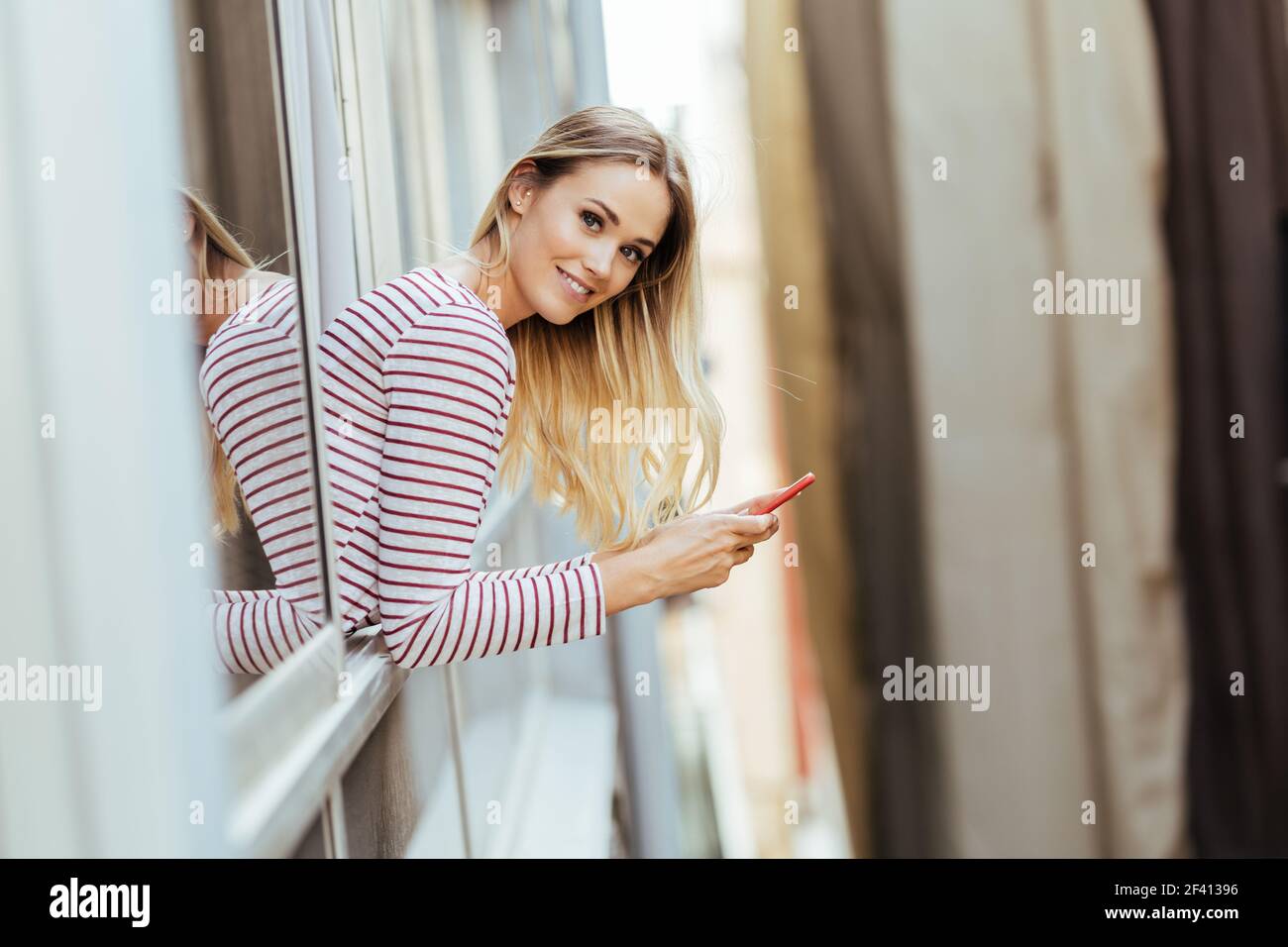 Young blonde woman leaning out of her house window using a smart phone ...