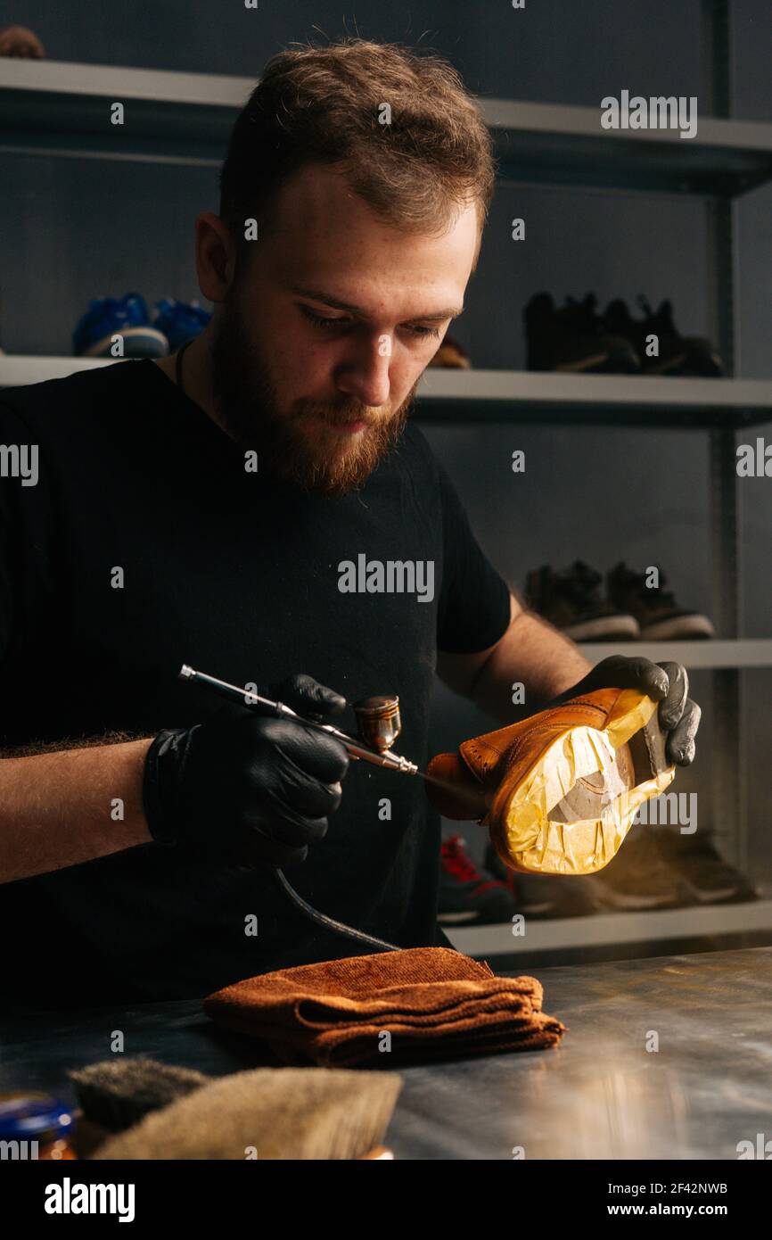 Portrait of bearded shoemaker wearing black gloves spraying paint of ...
