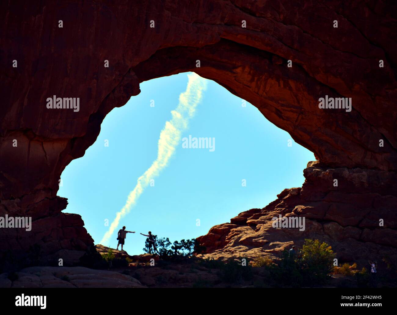A couple reaches with extended arms underneath a large natural stone ...