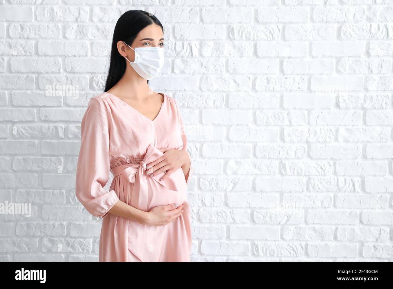 Young pregnant woman wearing medical mask on white brick background ...