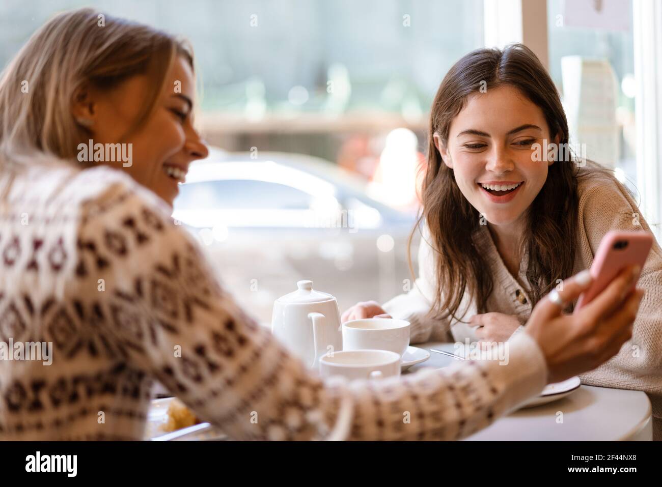 Two smiling young girlfriends using mobile phone while having lunch in ...