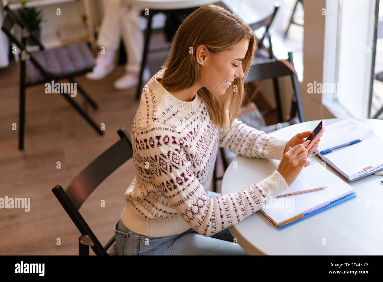 Smiling charming student girl using mobile phone while doing homework ...