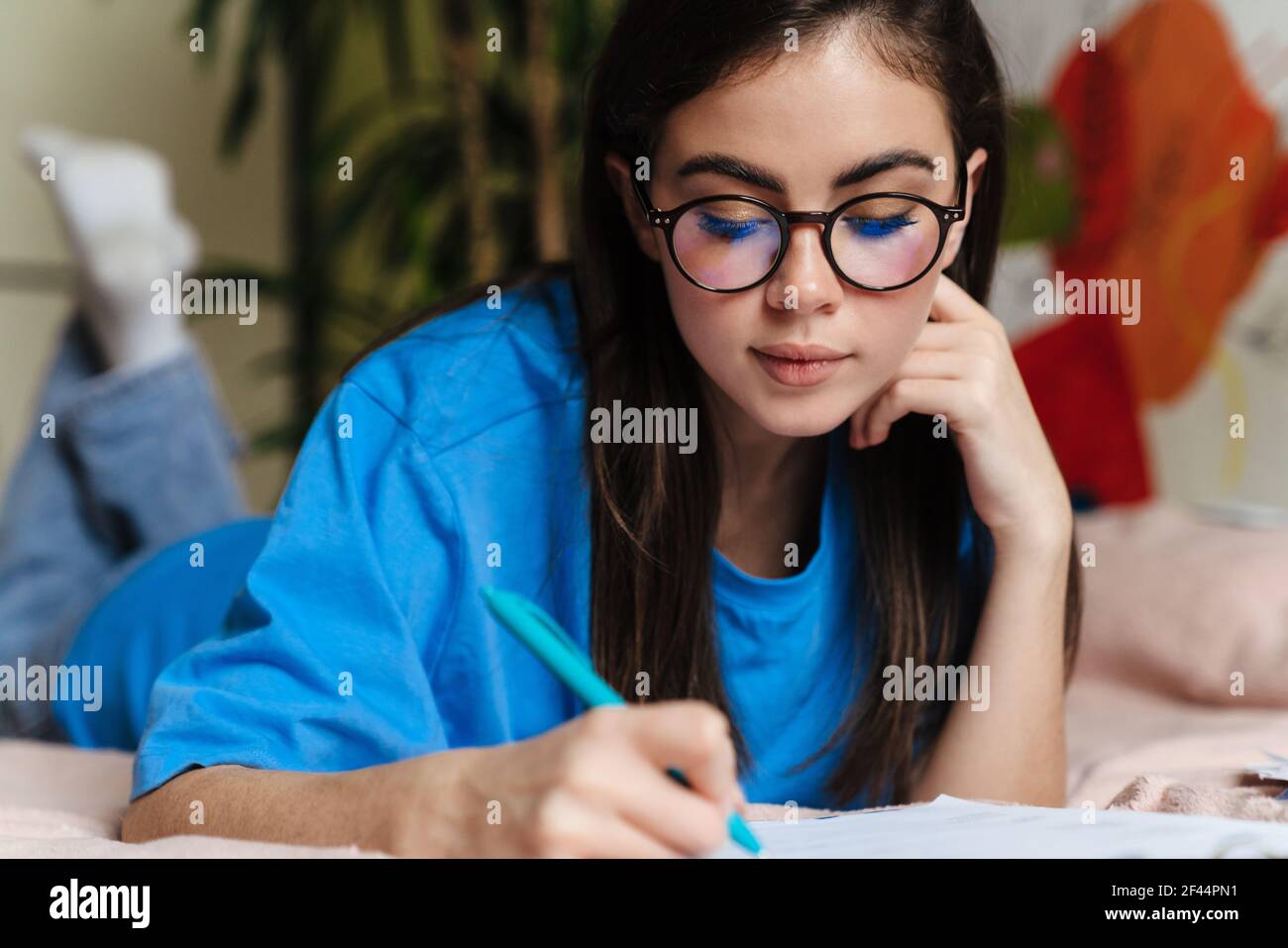 Focused brunette girl in eyeglasses doing homework while lying on bed ...