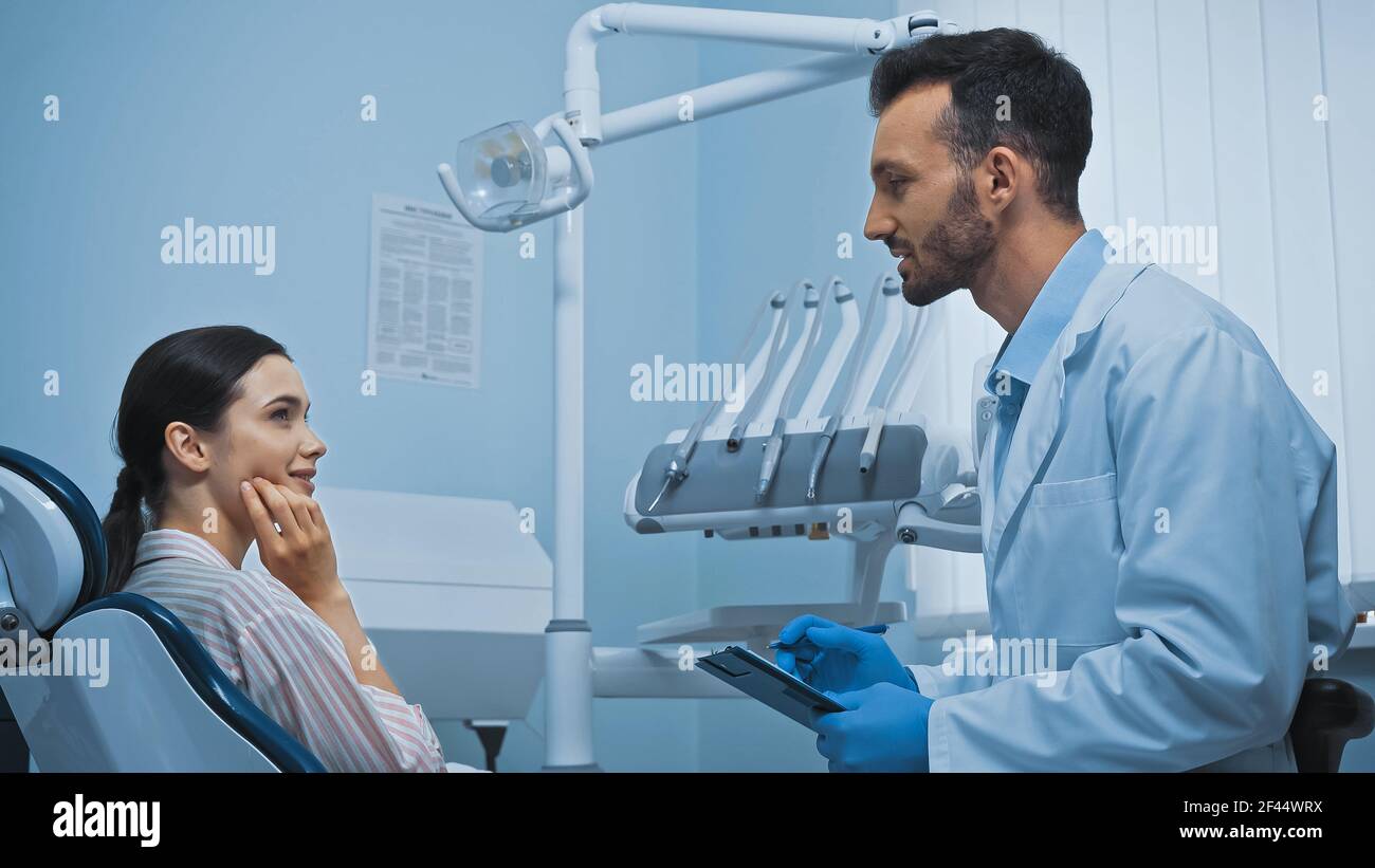 side view of dentist with clipboard near woman pointing at cheek in ...