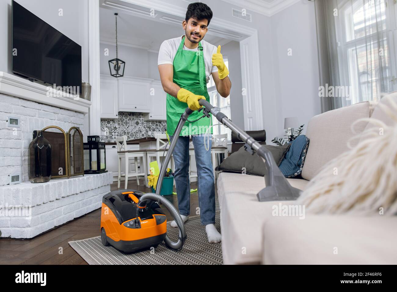 Indian young man cleaner in uniform cleaning sofa with modern vacuum ...