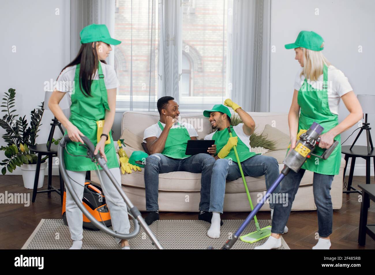 Male mixed race cleaners in green apron sitting on couch and playing on ...