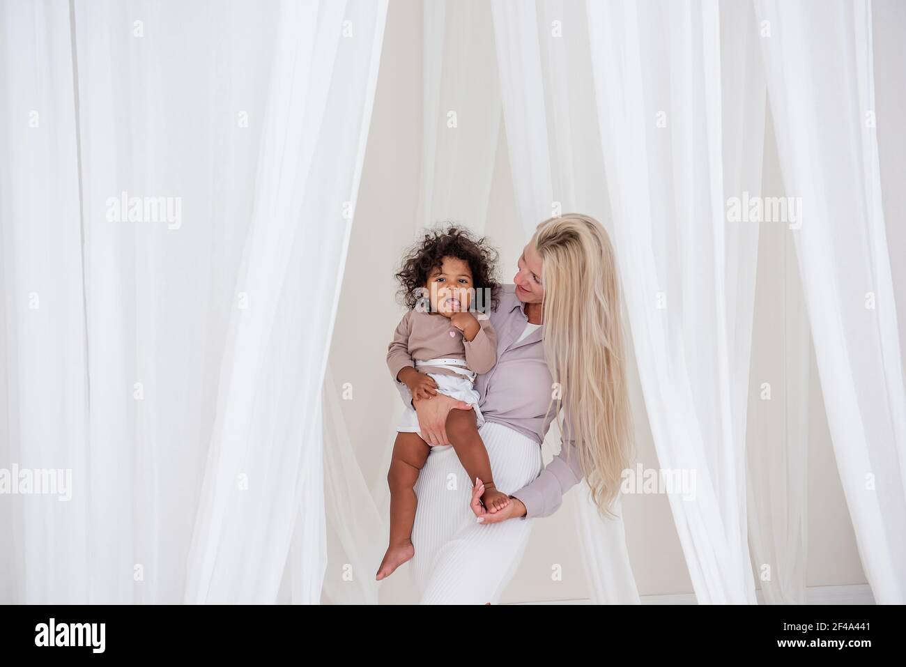 Young blond Caucasian mother is holding African American curly daughter ...