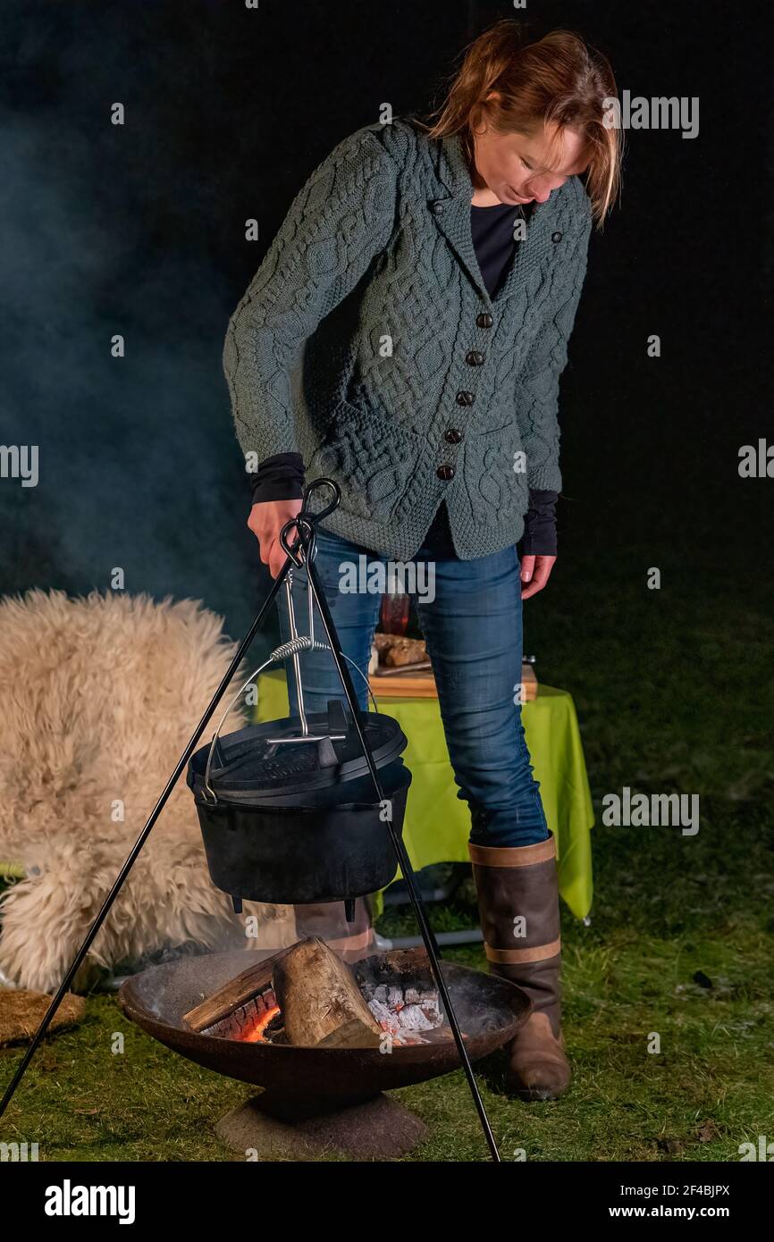 Young blond woman cooks dinner in a black kettle, outside on a winter ...