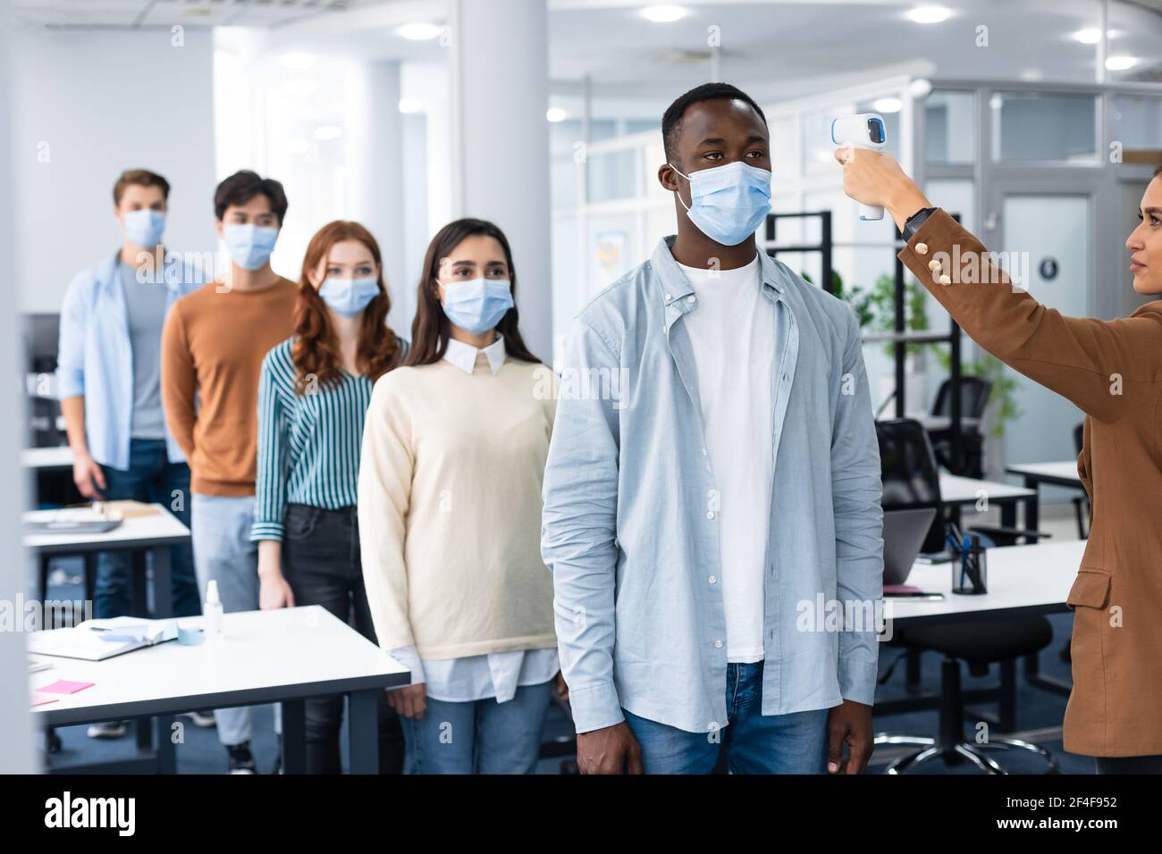 Woman with gun thermometer cheking diverse people temperature in line ...