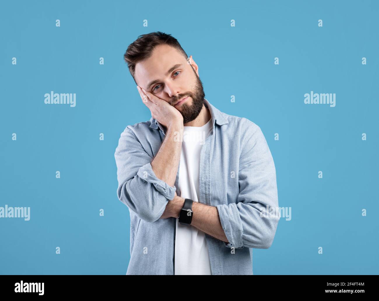 Bored young guy leaning on his hand and looking at camera over blue ...