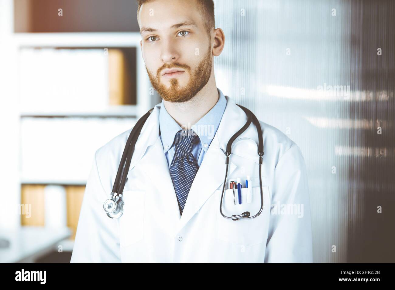 Red-bearded doctor standing straight in sunny clinic near his working ...