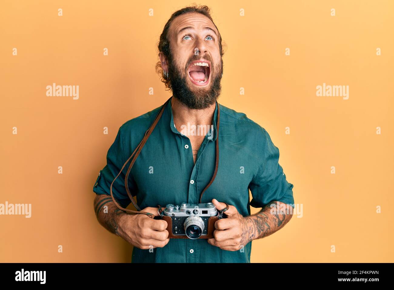 Handsome man with beard and long hair holding vintage camera angry and ...