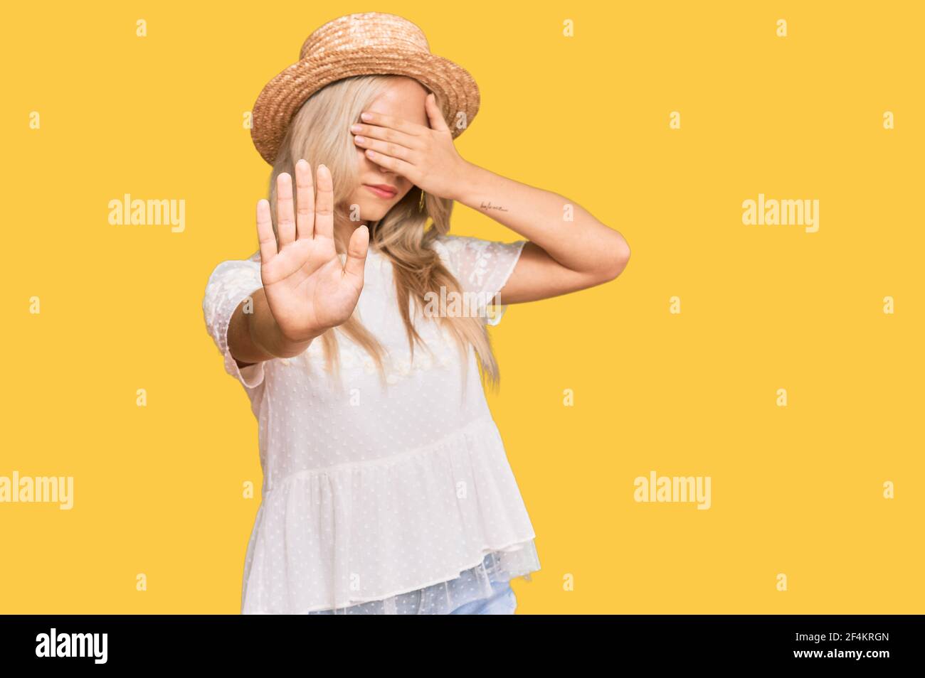 Young blonde girl wearing summer hat covering eyes with hands and doing ...