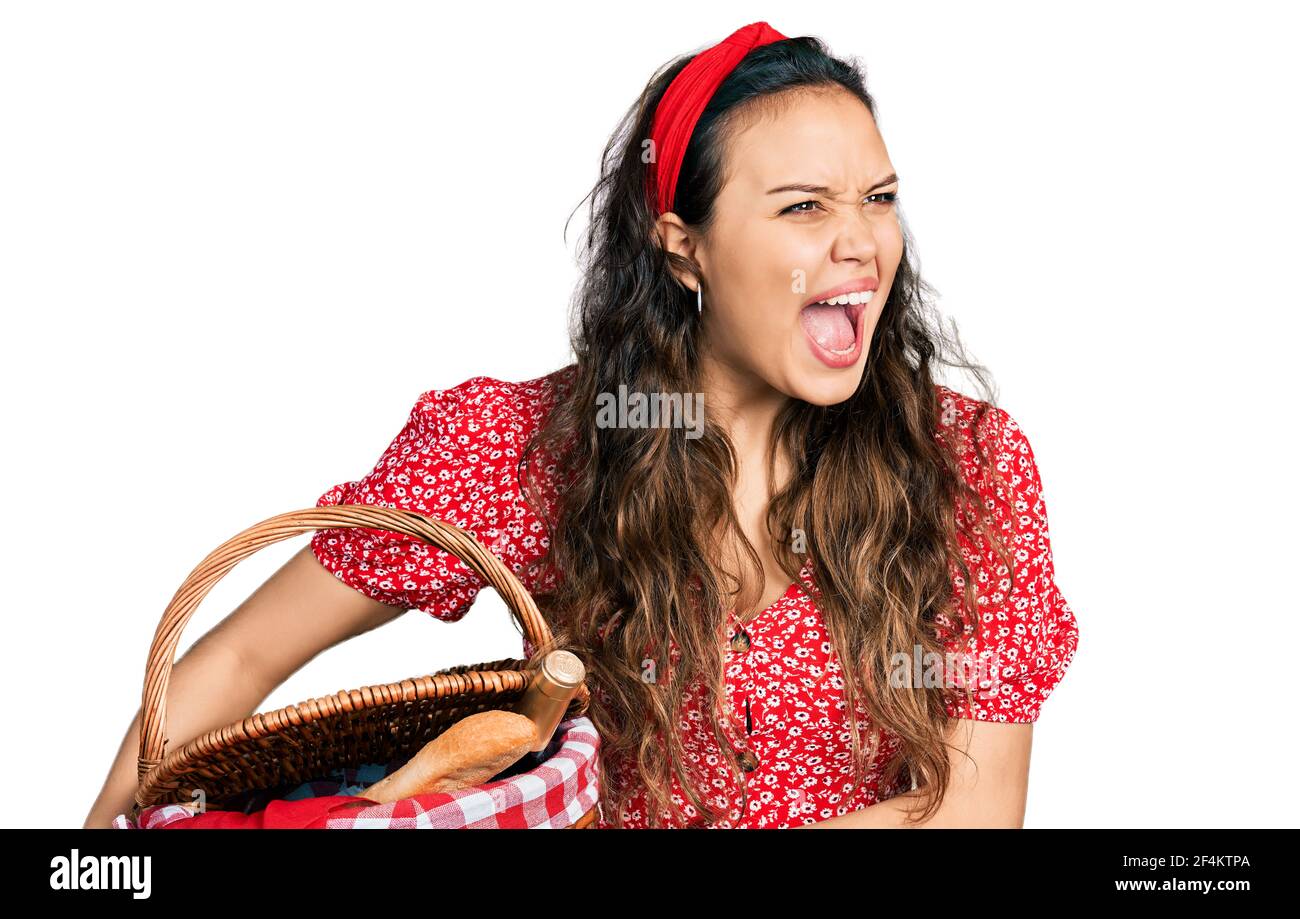 Young hispanic girl holding picnic wicker basket with bread angry and ...