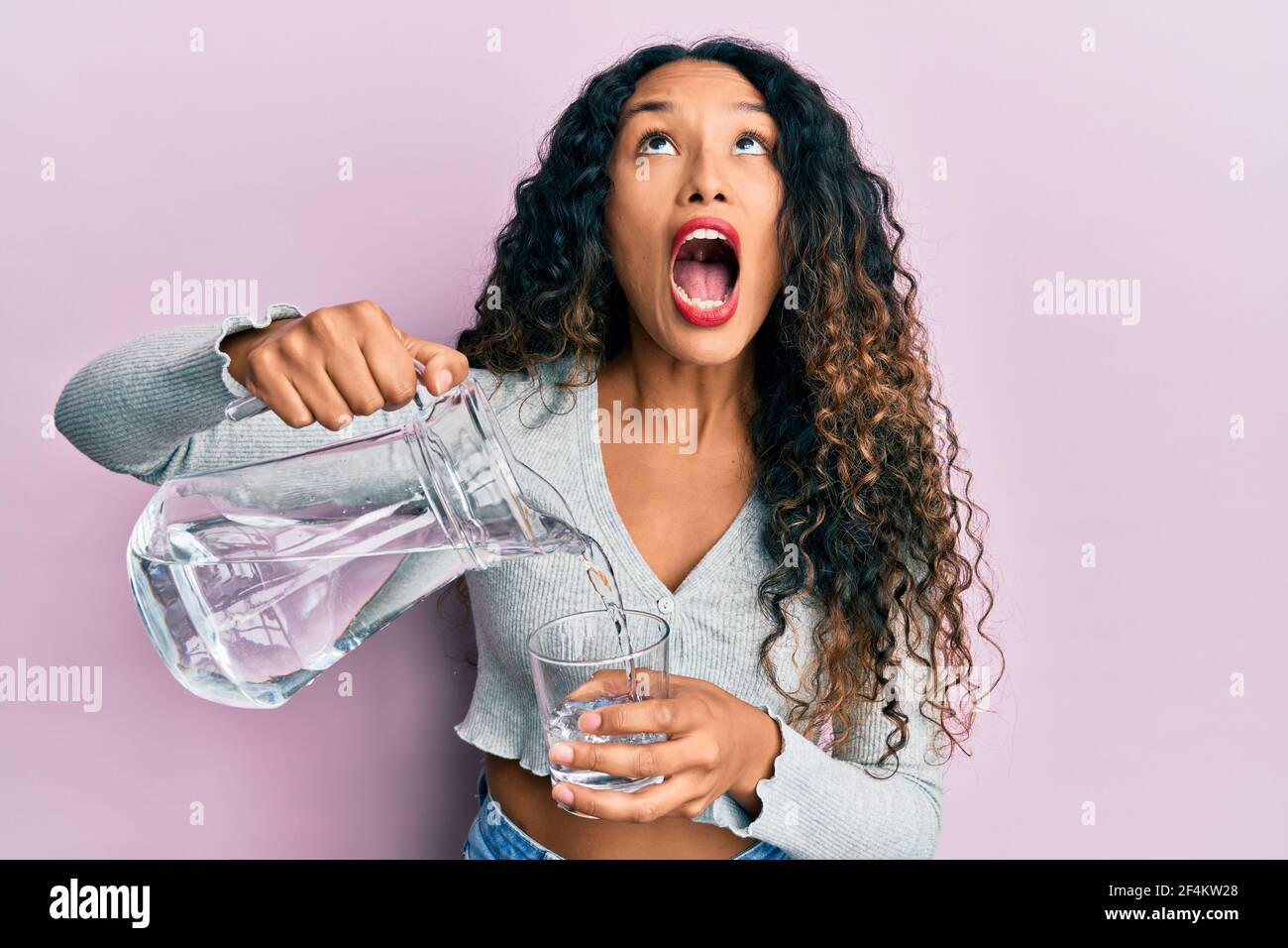 Young latin woman pouring water angry and mad screaming frustrated and ...