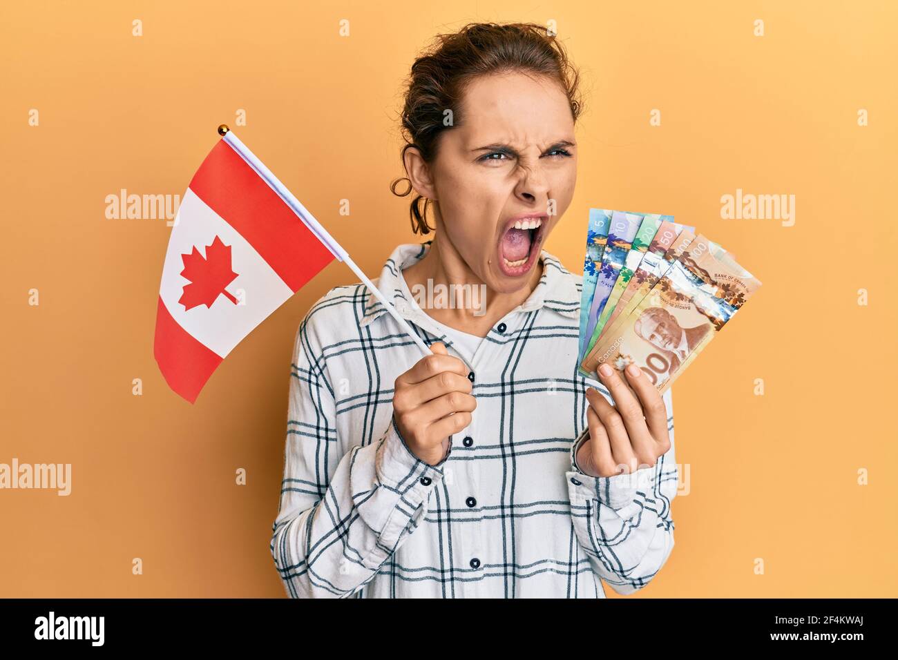 Young brunette woman holding canada flag and dollars angry and mad ...