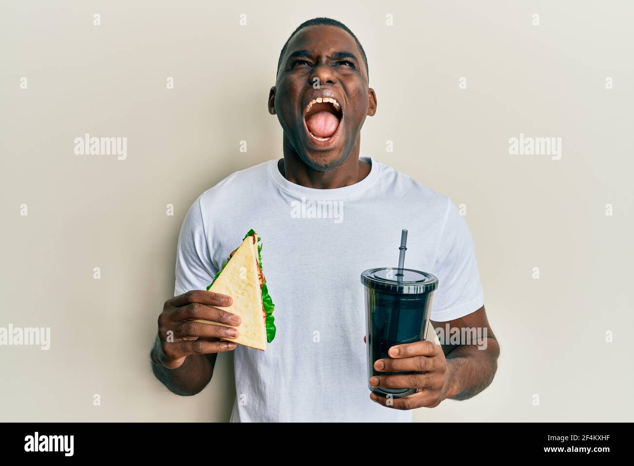 Young african american man eating sandwich and drinking soda angry and ...