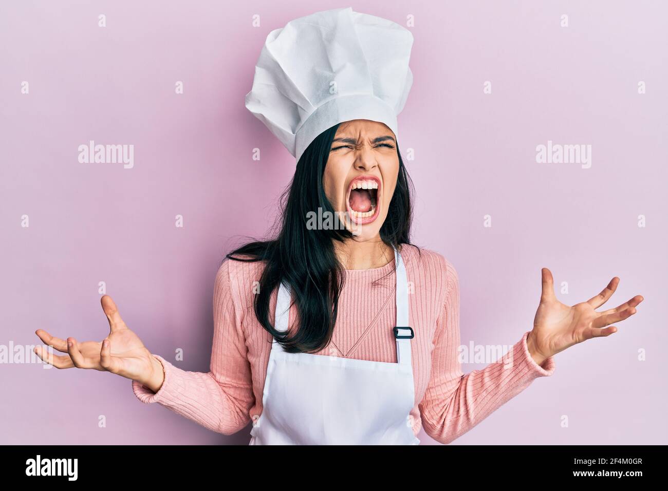 Young hispanic woman wearing baker uniform and cook hat crazy and mad ...