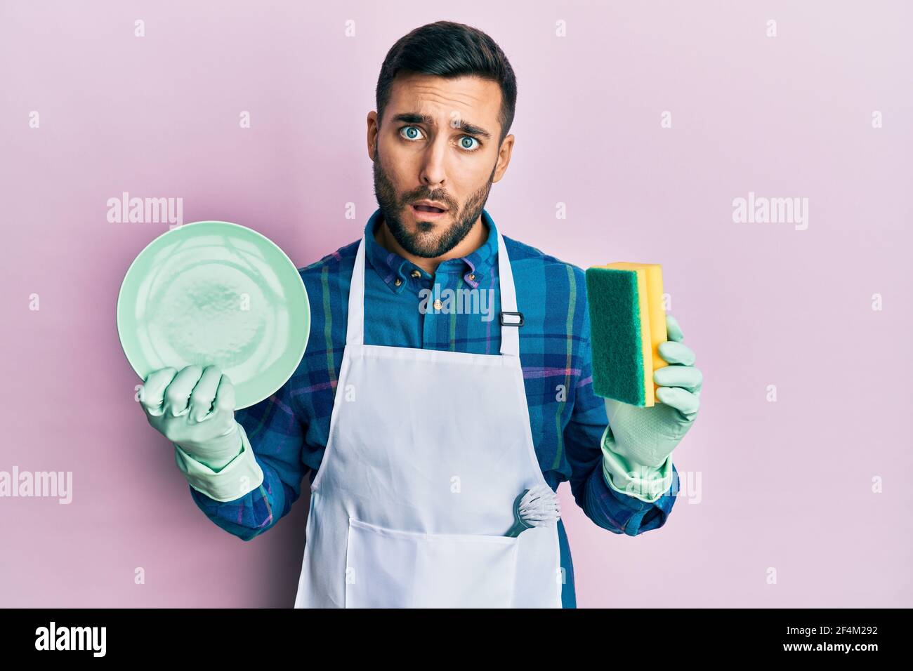 Young hispanic man wearing apron holding scourer washing dishes in ...