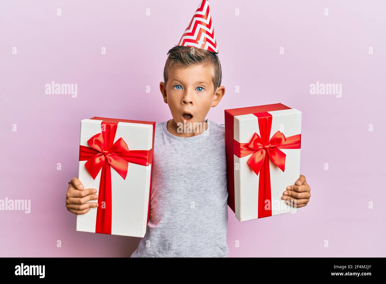 Adorable caucasian kid wearing birthday hat holding presents afraid and ...
