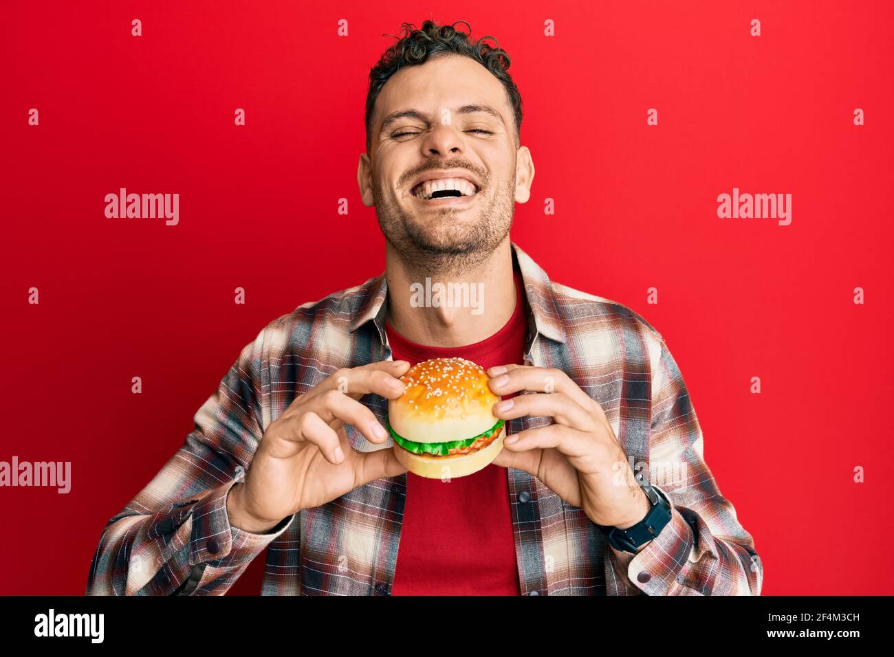 Young hispanic man eating a tasty classic burger smiling and laughing ...