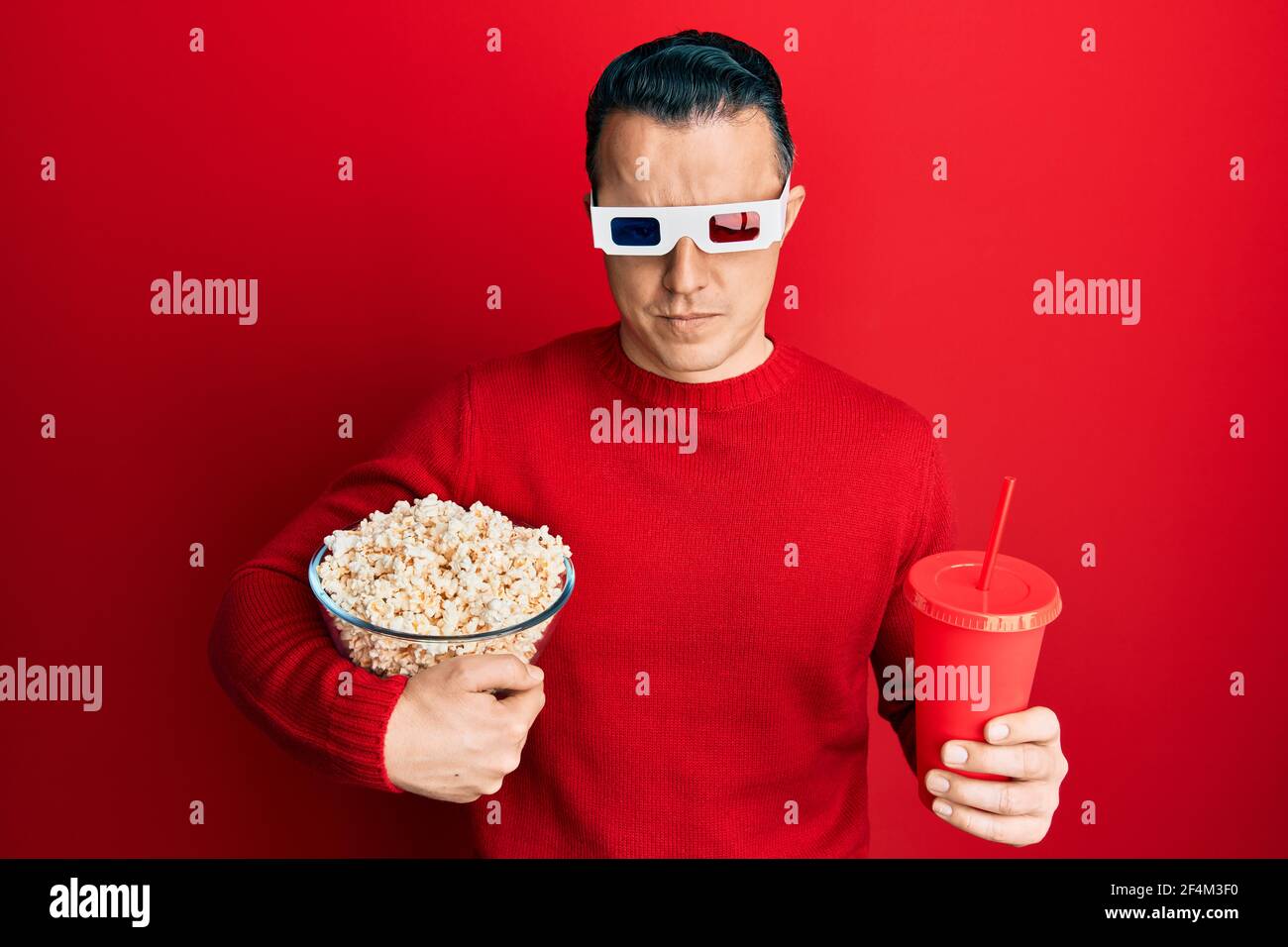 Handsome young man eating popcorn and soda wearing 3d glasses skeptic ...