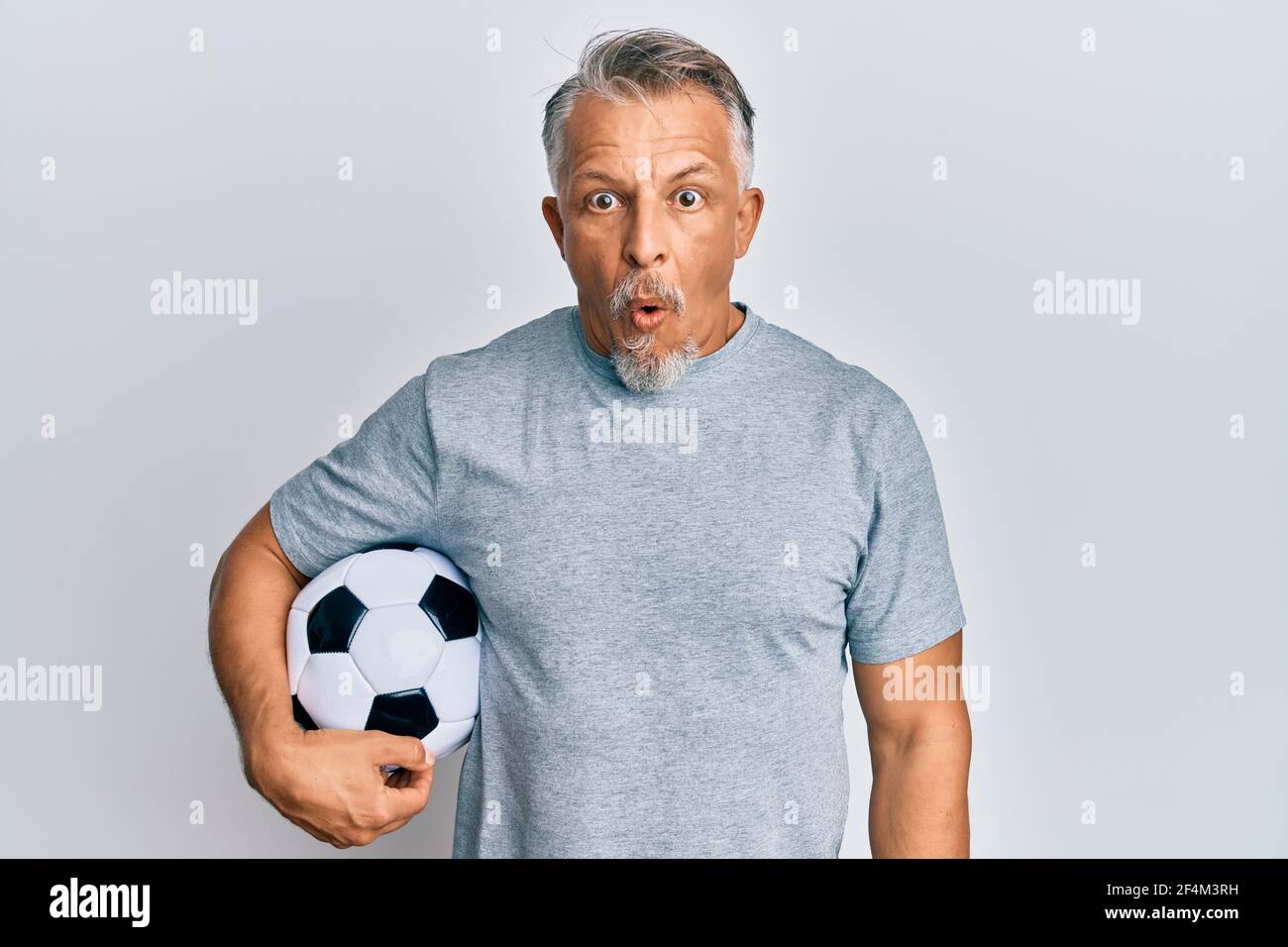 Middle age grey-haired man holding soccer ball scared and amazed with ...