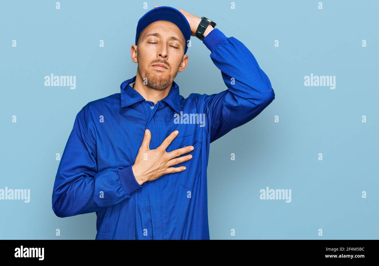 Bald man with beard wearing builder jumpsuit uniform touching forehead ...