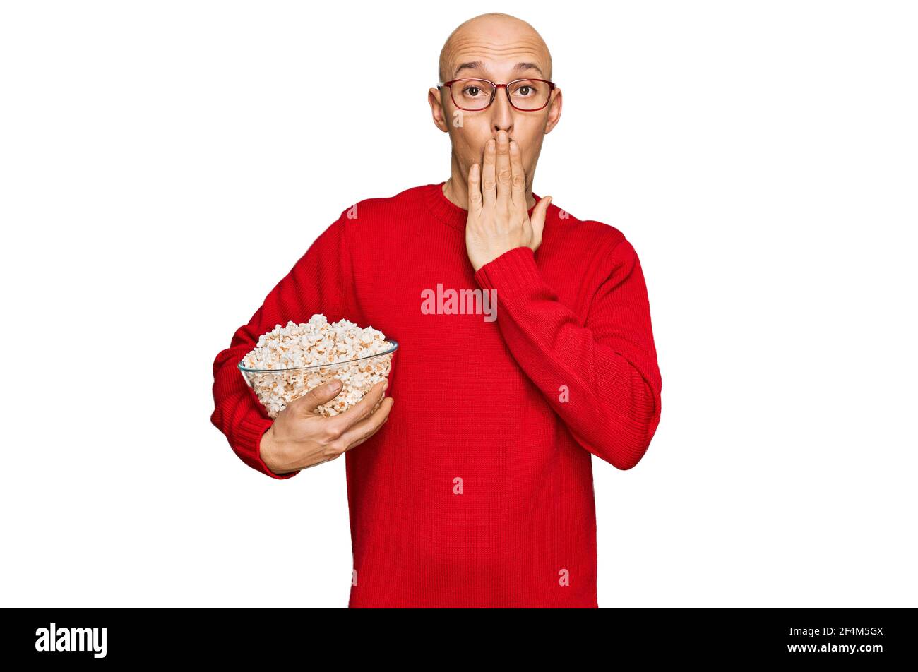 Bald man with beard eating popcorn covering mouth with hand, shocked ...
