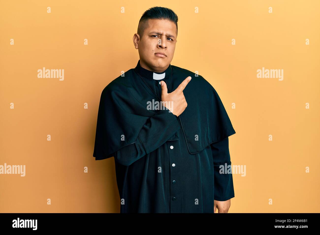Young latin priest man standing over yellow background pointing with ...