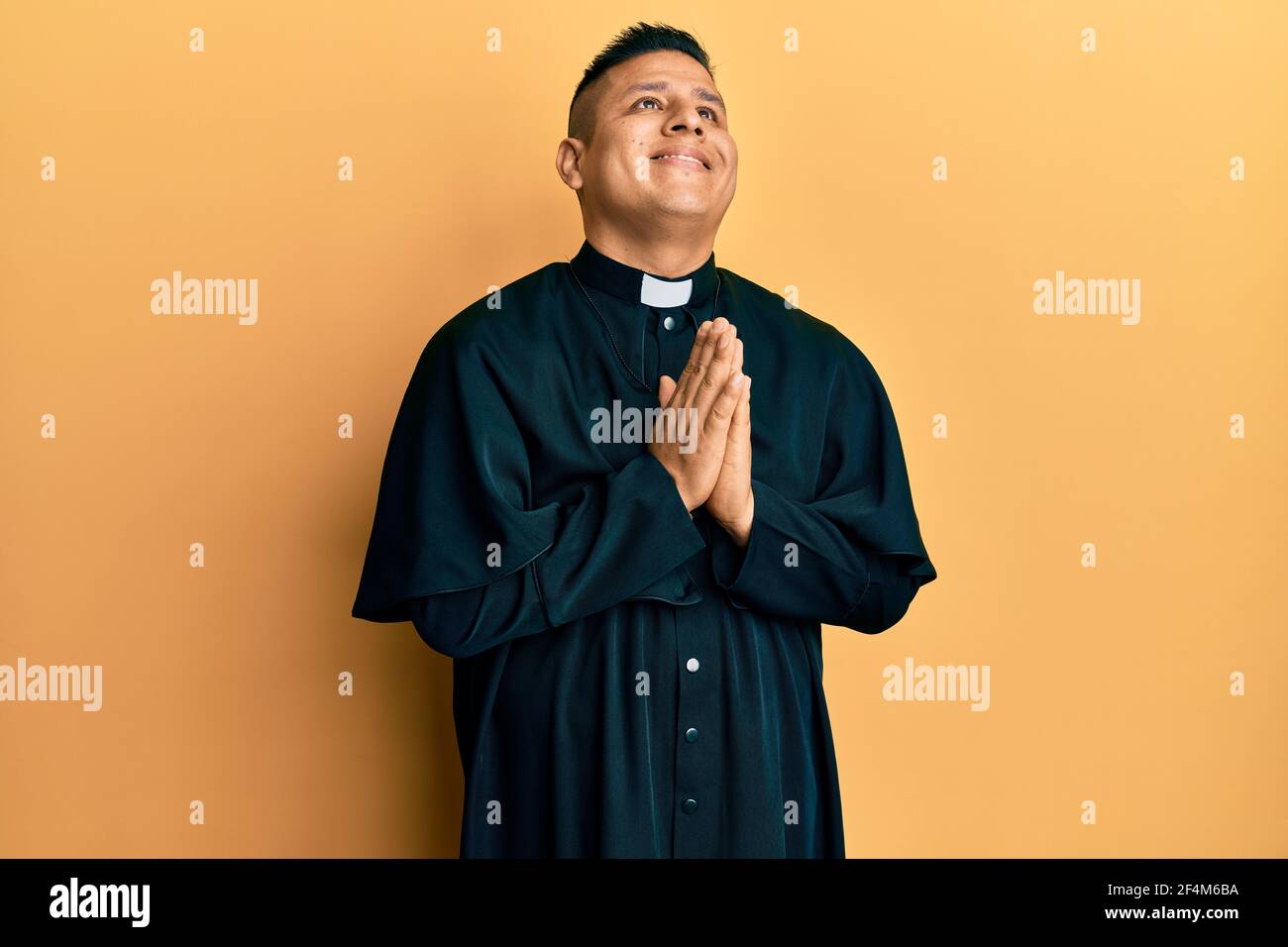Young latin priest man standing over yellow background begging and ...