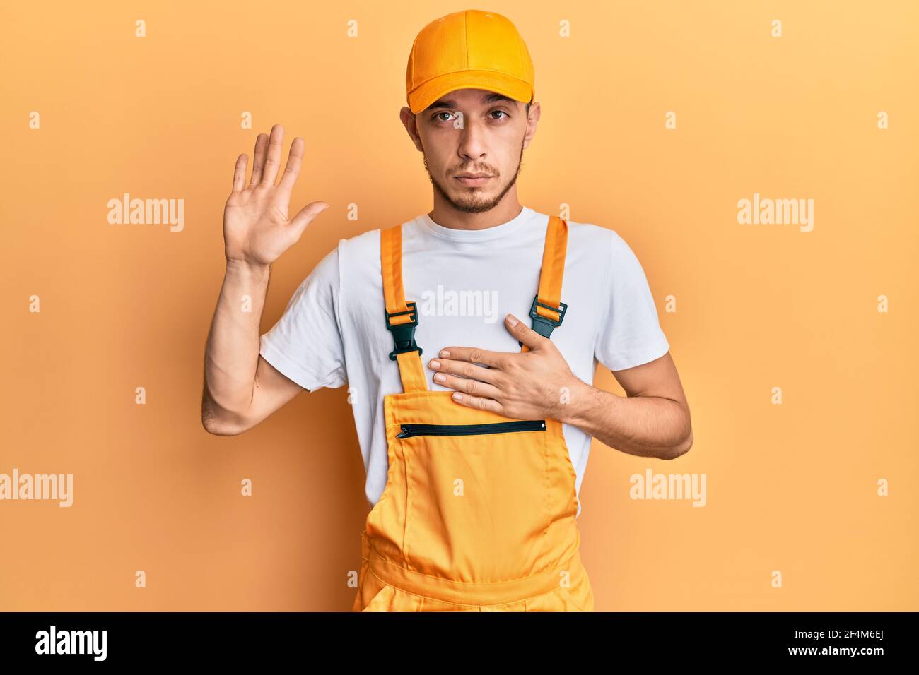 Hispanic young man wearing handyman uniform swearing with hand on chest ...