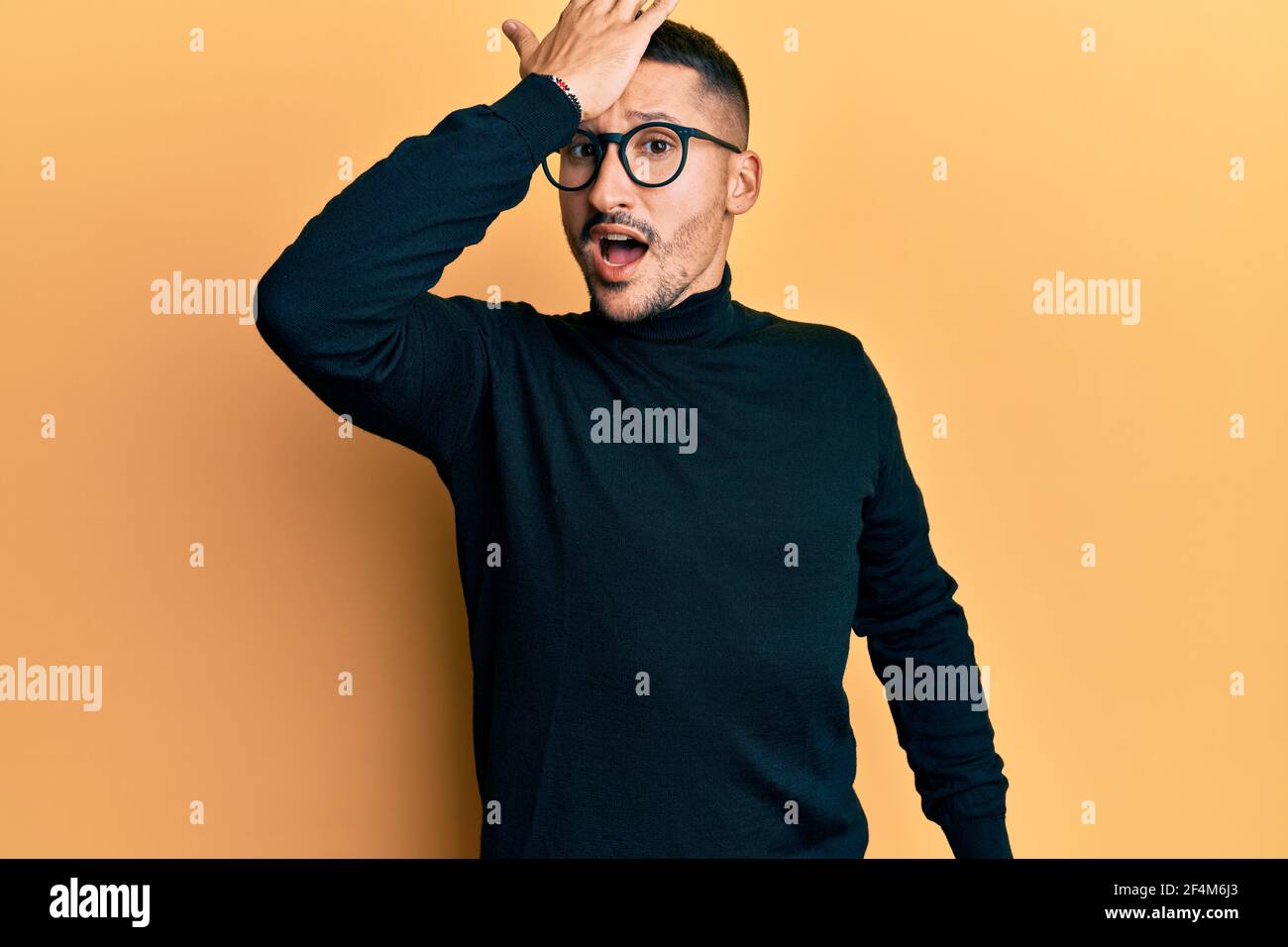 Handsome man with tattoos wearing turtleneck sweater and glasses surprised with hand on head for ...