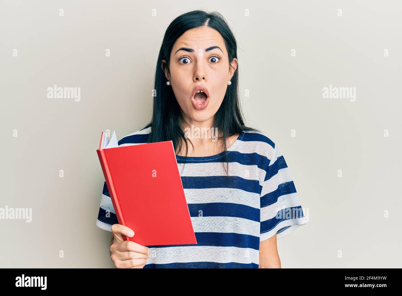 Beautiful young woman reading book scared and amazed with open mouth ...