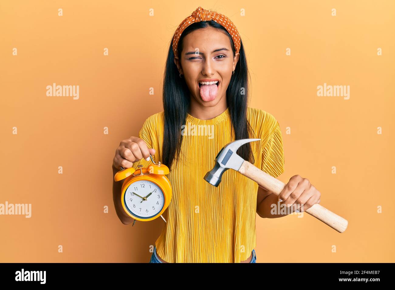 Young Brunette Woman Holding Alarm Clock And Hammer Sticking Tongue Out