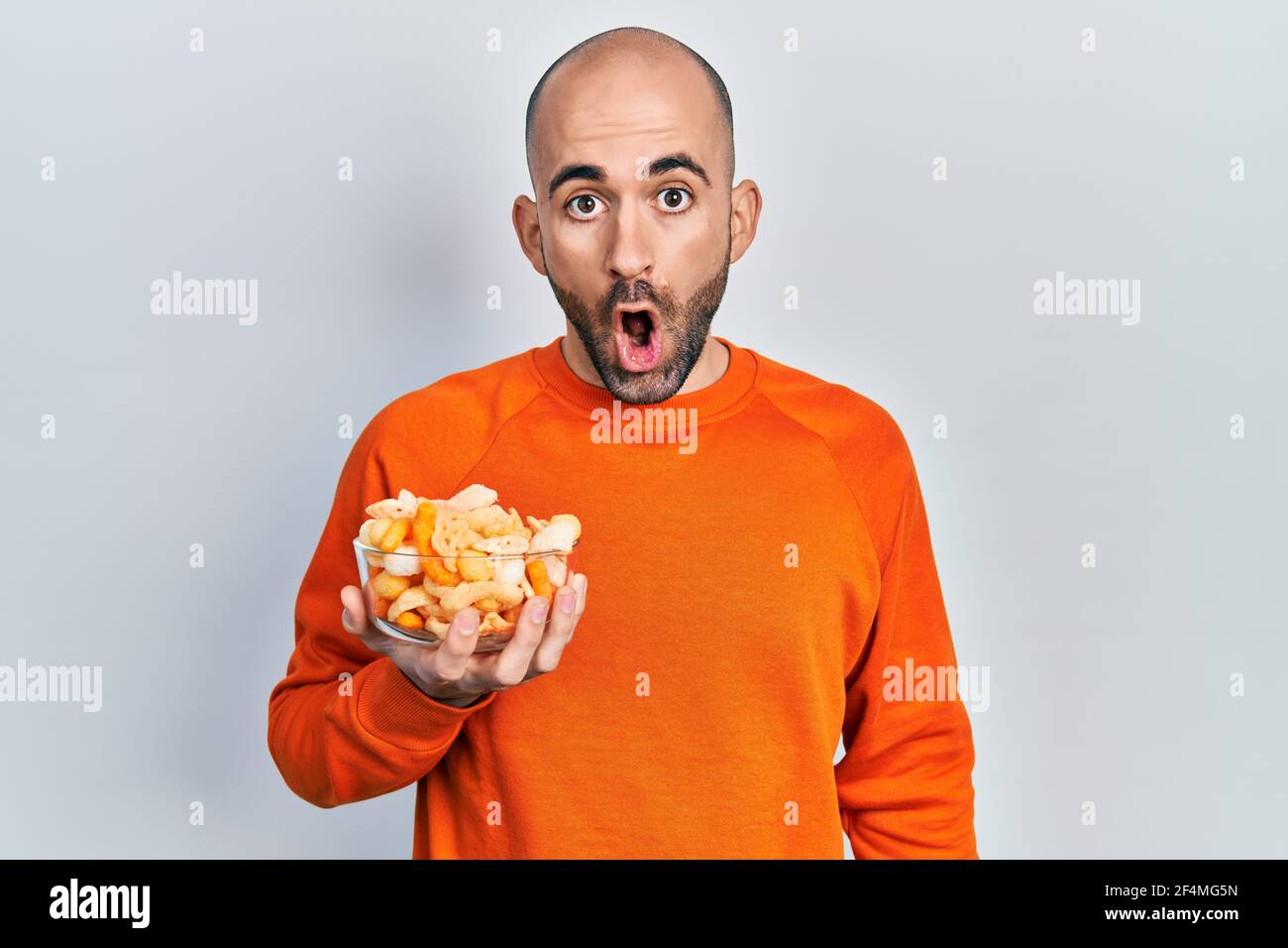 Young bald man holding potato chip scared and amazed with open mouth ...