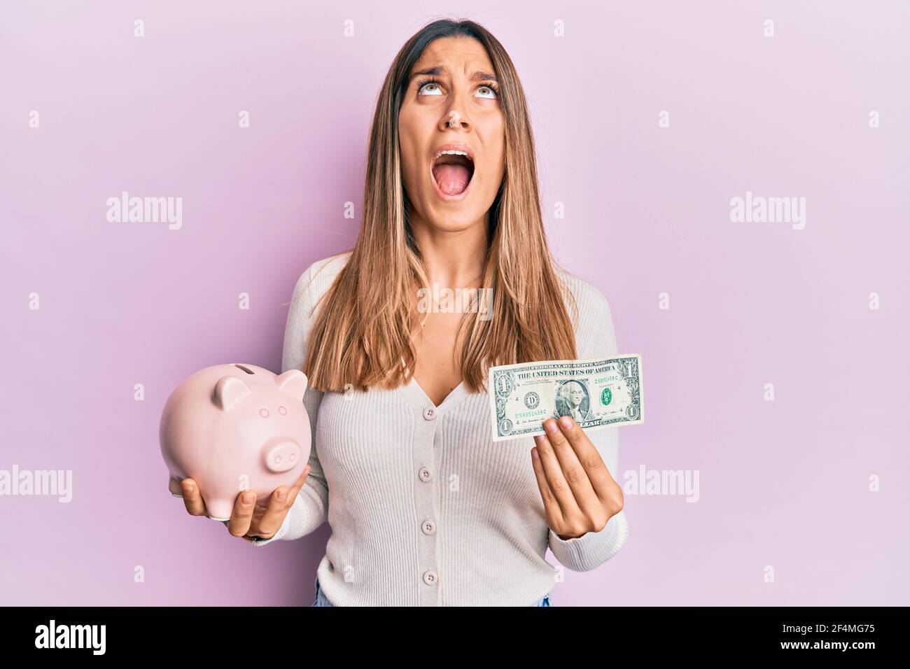 Brunette young woman holding one dollar banknote and piggy bank angry ...
