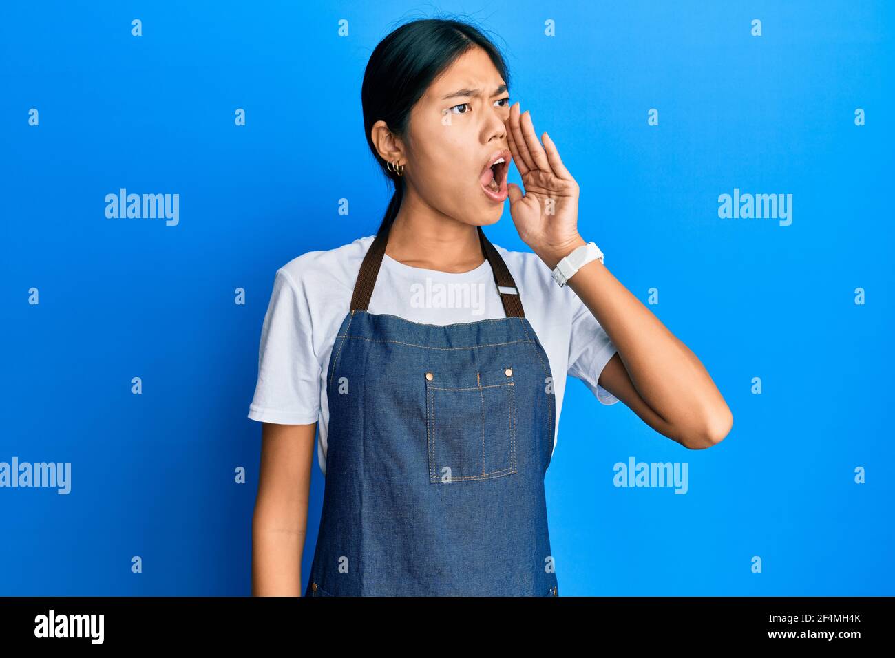 Young chinese woman wearing waiter apron shouting and screaming loud to ...