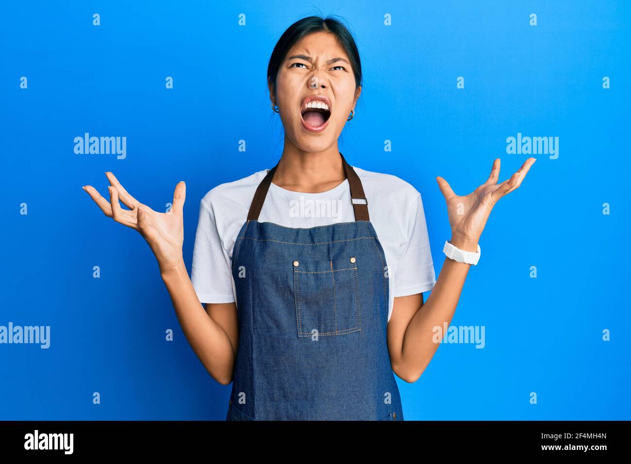Young chinese woman wearing waiter apron crazy and mad shouting and ...