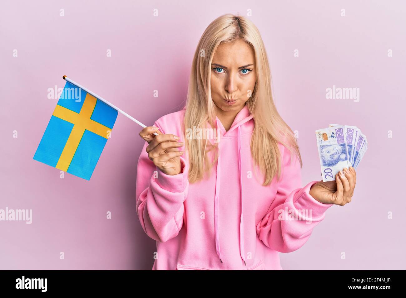 Young blonde woman holding sweden flag and krone banknotes skeptic and ...